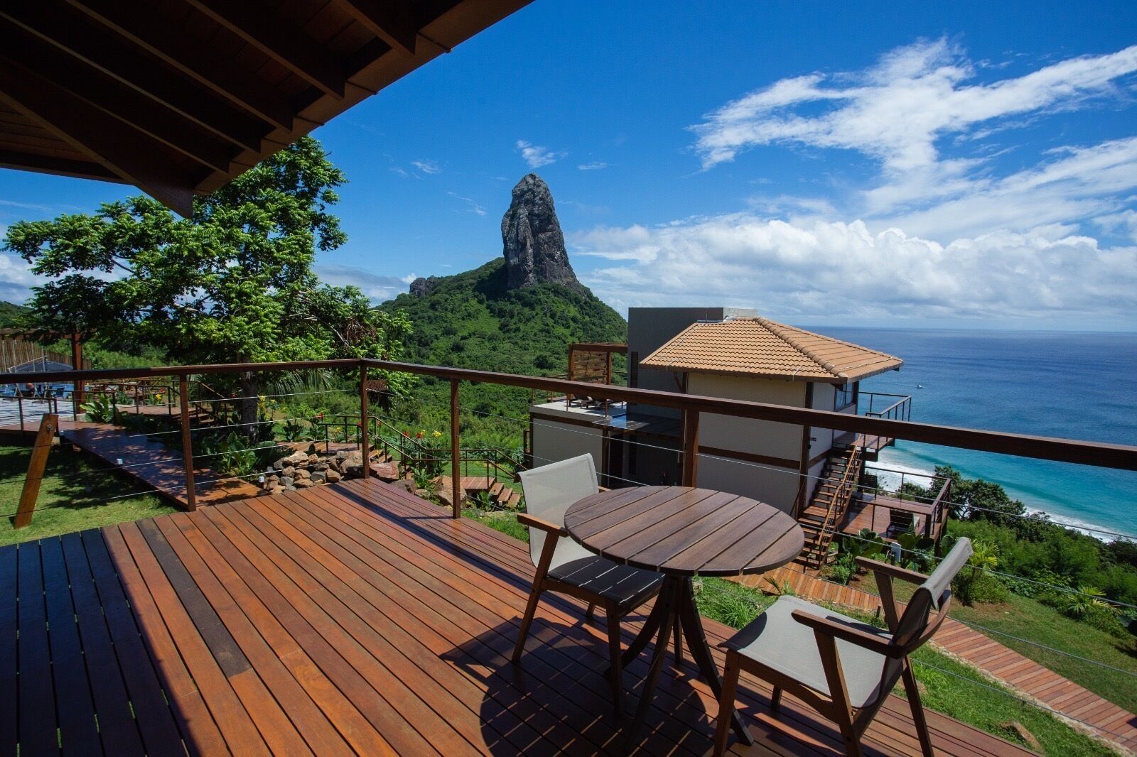 Um deck de madeira com mesa e cadeiras, com vista para a costa e uma montanha ao longe.