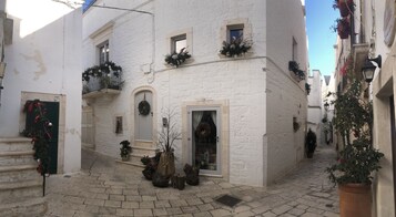 Uma rua estreita com um edifício branco, apresentando uma pequena varanda com plantas em vasos e uma porta com grinalda.