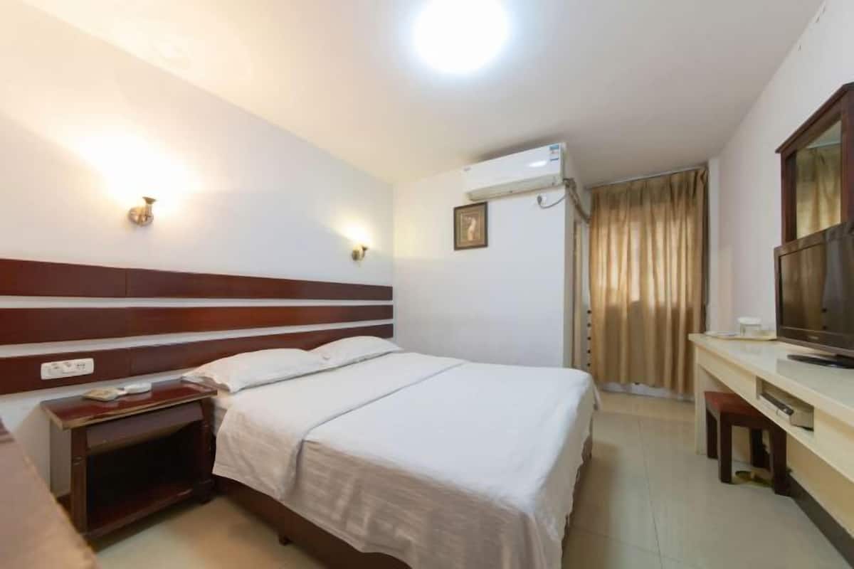 Quarto de hotel com cama, mesa de cabeceira, luminária, aparelho de ar condicionado, janela com cortinas e televisão.