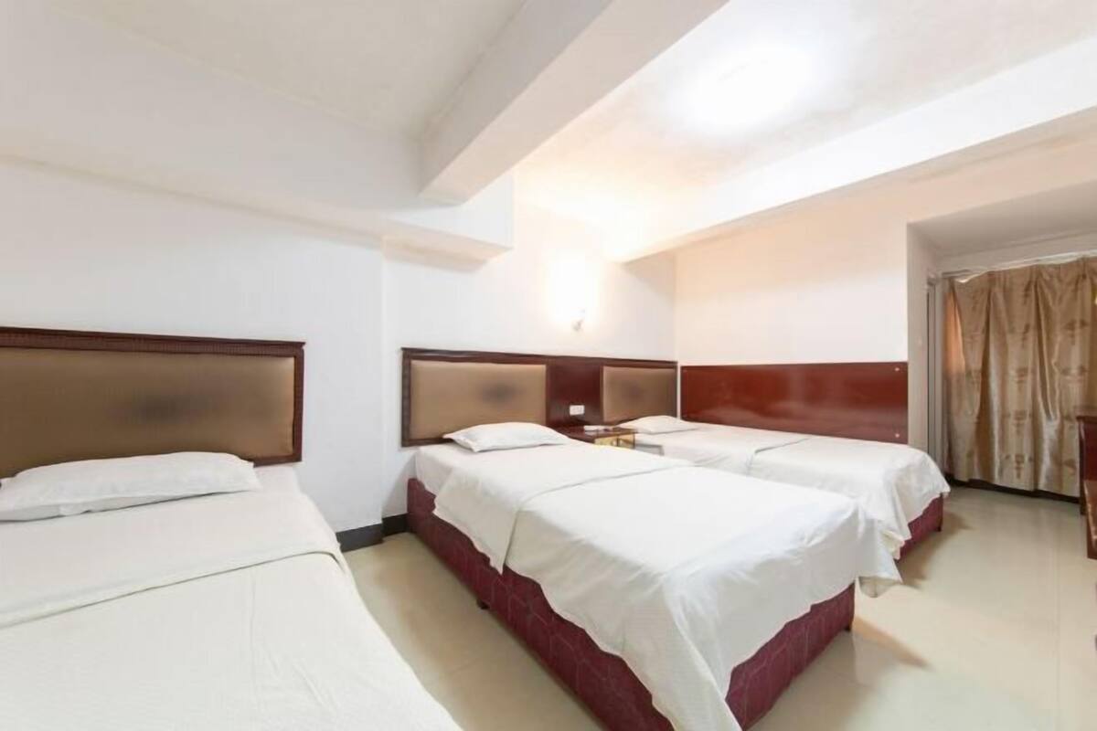 Quarto de hotel com três camas, cabeceira de madeira, mesa de cabeceira e janela com cortinas.