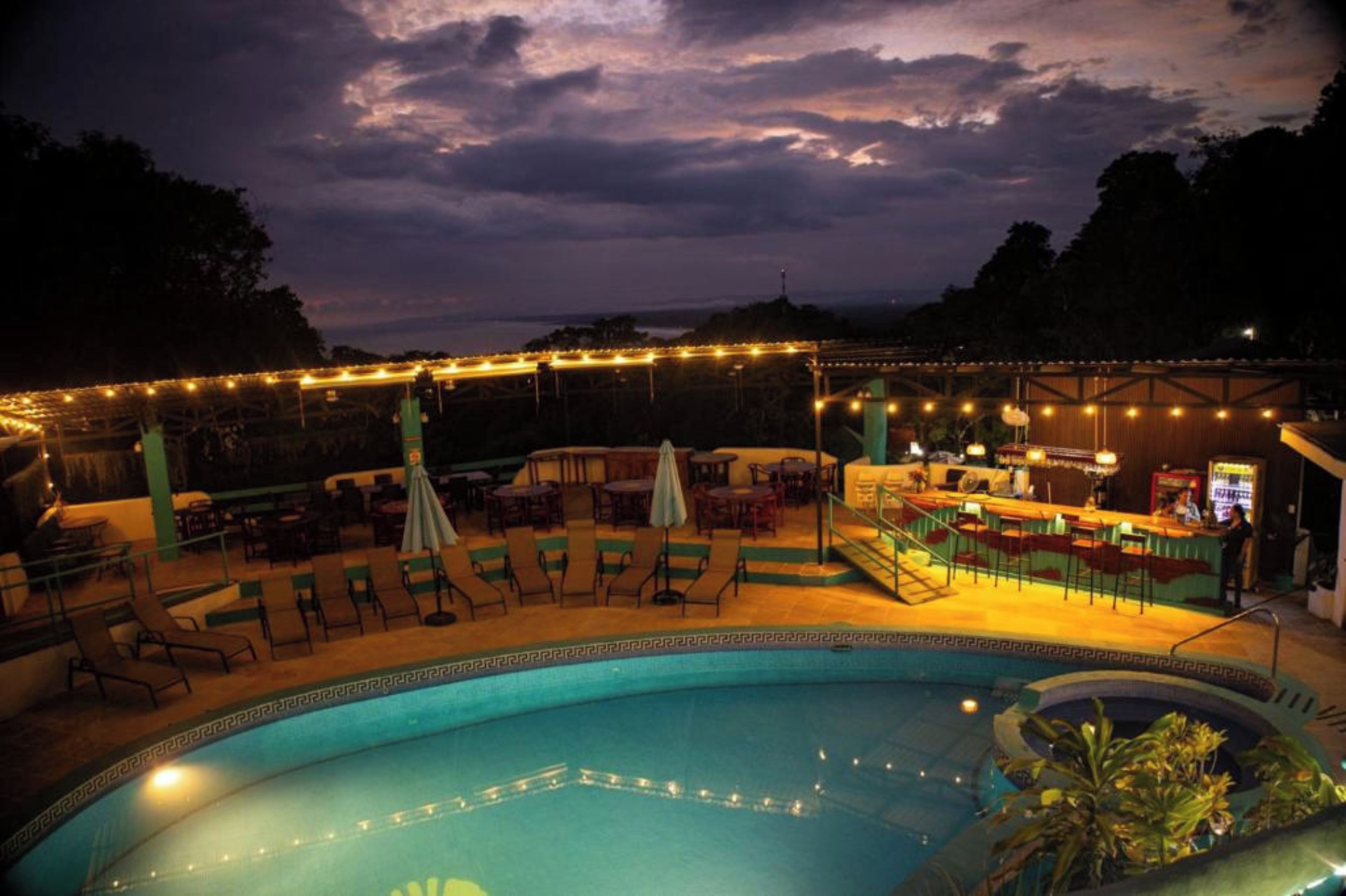 Piscina ao ar livre com espreguiçadeiras e guarda-sóis, cercada por área coberta com mesas e cadeiras, e um bar com vista para o céu.