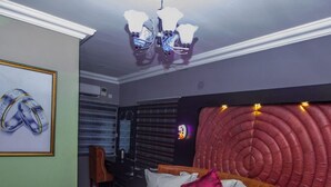 Quarto com cama, almofadas, manta, cabeceira, luminária no teto, quadro na parede e janela com persianas.