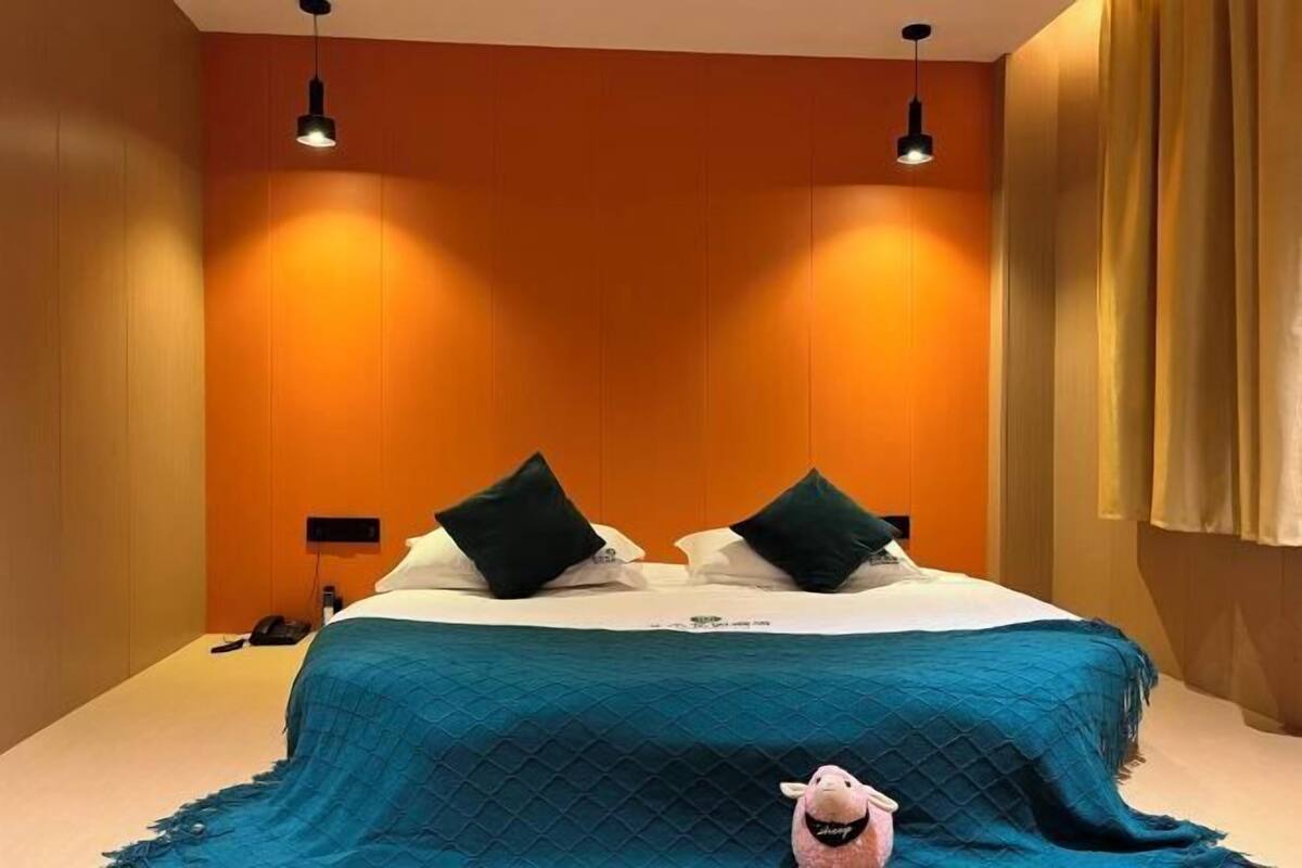 Quarto de hotel com uma cama, duas almofadas pretas, um cobertor azul, um brinquedo de pelúcia rosa e duas luminárias pendentes.