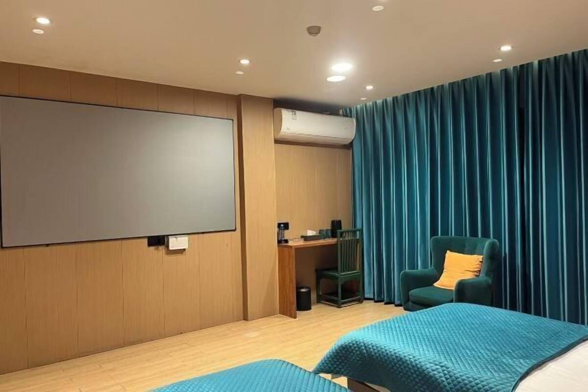 Quarto de hotel com uma cama grande, uma escrivaninha com cadeira, aparelho de ar condicionado e uma janela ampla com cortinas.
