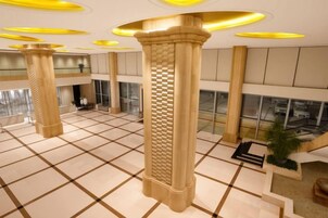 Um hall espaçoso com grandes colunas, piso xadrez e vista para o estacionamento através de amplas janelas.