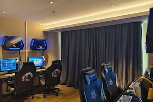Sala de jogos com vários monitores, cadeiras gamer e uma grande janela com cortinas.