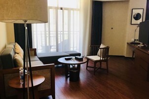 Quarto de hotel com sofá, mesa redonda pequena, cadeira, luminária, televisão e janela com cortinas.