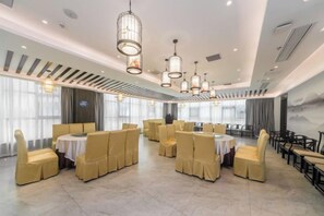 Uma sala de banquetes com mesas redondas e cadeiras, teto alto com luminárias pendentes e grandes janelas.