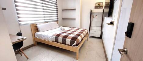 Quarto com cama de madeira, escrivaninha, cadeira, guarda-roupa e televisão na parede.