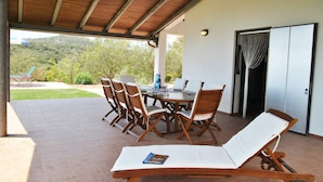 Um pátio externo com mesa e cadeiras, uma poltrona de descanso e vista para uma paisagem verde.