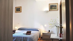 Quarto com duas camas, uma mesinha de cabeceira com abajur, um vaso com flores e duas imagens emolduradas na parede.