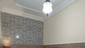 Quarto com parede estampada, cama com colcha branca e cinza, cabeceira de madeira, mesa de cabeceira com abajur e uma pequena estante.