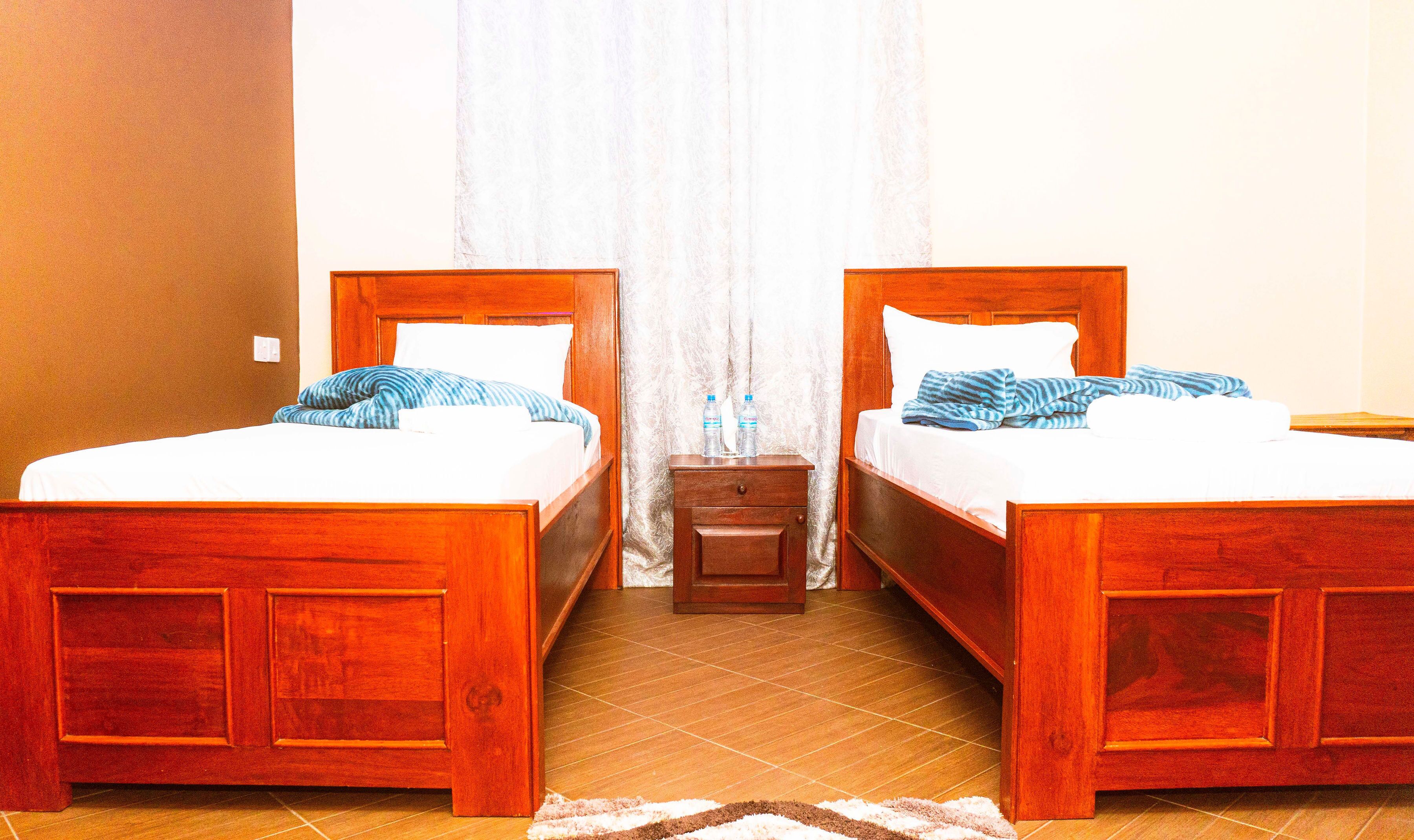 Quarto com duas camas de madeira, uma pequena mesa de cabeceira de madeira e uma cortina branca.