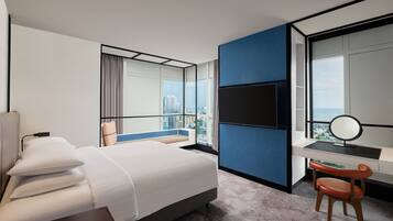 Quarto de hotel moderno com uma cama grande, uma escrivaninha com cadeira, televisão e vista para a cidade.
