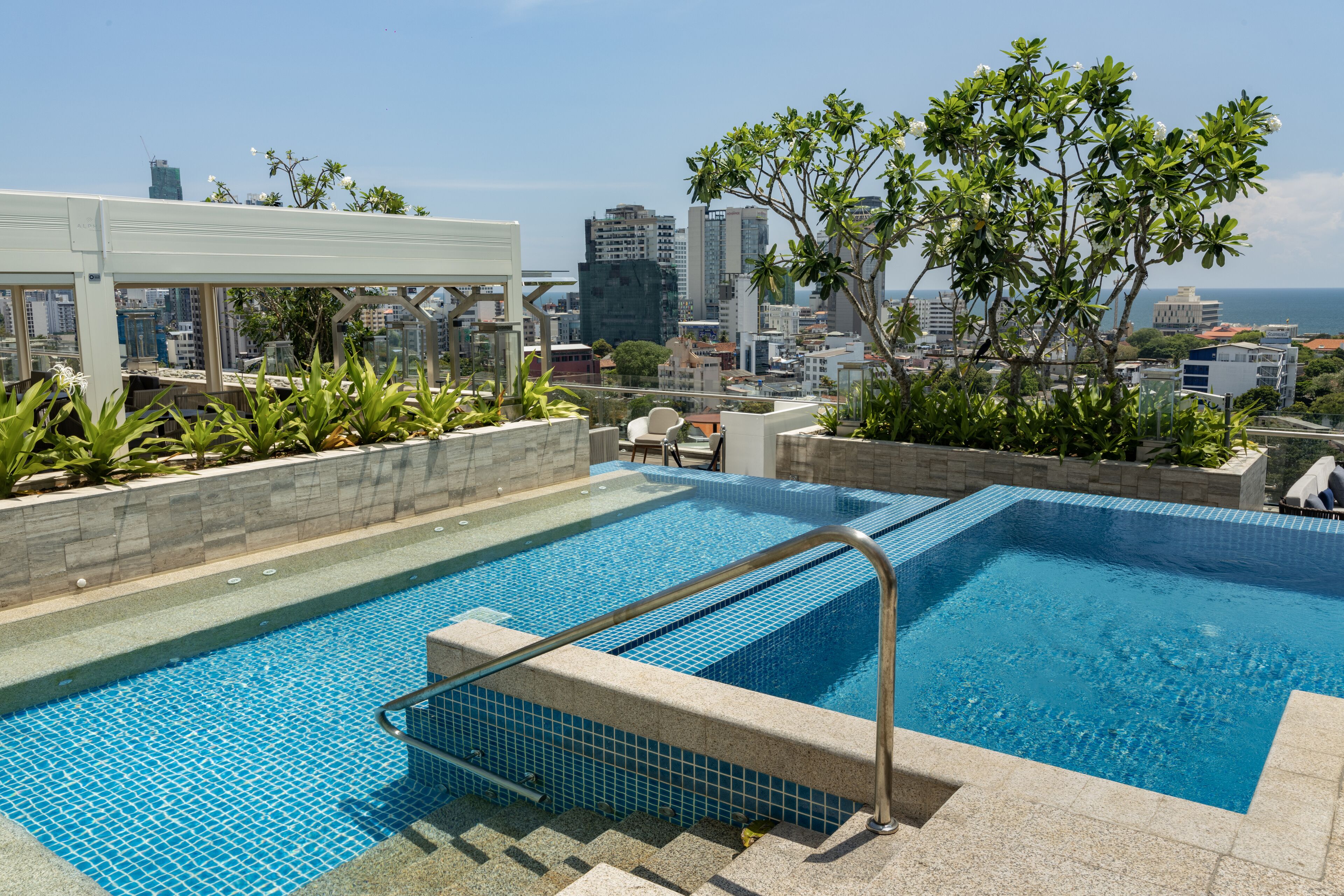 Piscina no terraço com vista para a cidade, cercada por plantas e uma pérgola.