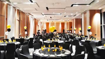 Uma sala de banquetes com mesas redondas preparadas para refeições, equipada com cadeiras, pratos, copos e suco de laranja.