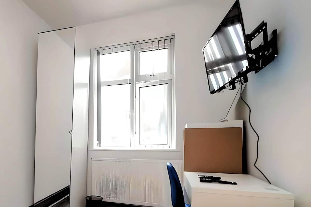 Quarto de hotel com cama, escrivaninha, cadeira, televisão fixada na parede e janela com cortinas.