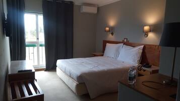 Quarto de hotel com cama, mesas de cabeceira, escrivaninha, cadeira, janela com cortinas e luminárias fixadas na parede.