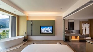Quarto de hotel moderno com uma cama grande, uma banheira, uma TV fixada na parede e vista para montanhas pela janela.