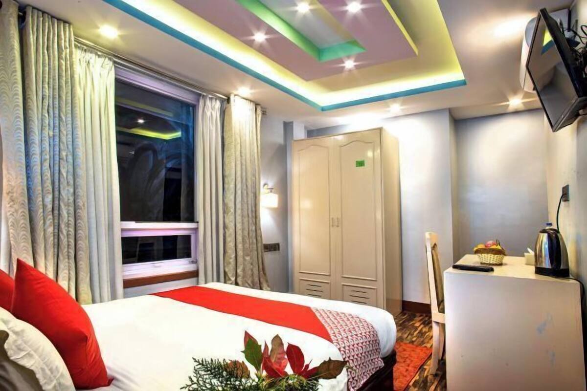 Quarto de hotel com cama, guarda-roupa, escrivaninha com chaleira e televisão.