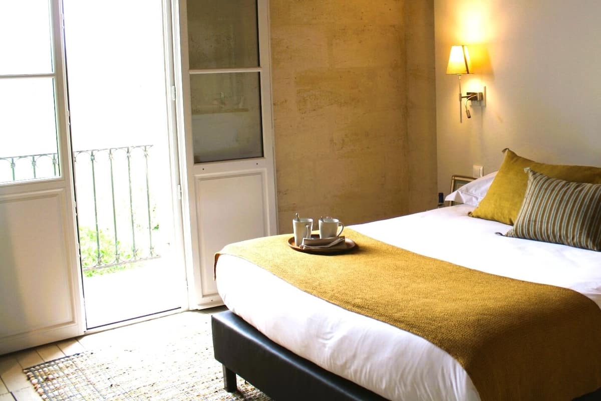 Quarto de hotel com uma cama, um suporte com xícaras e uma chaleira, uma janela com vista e duas luminárias de parede.