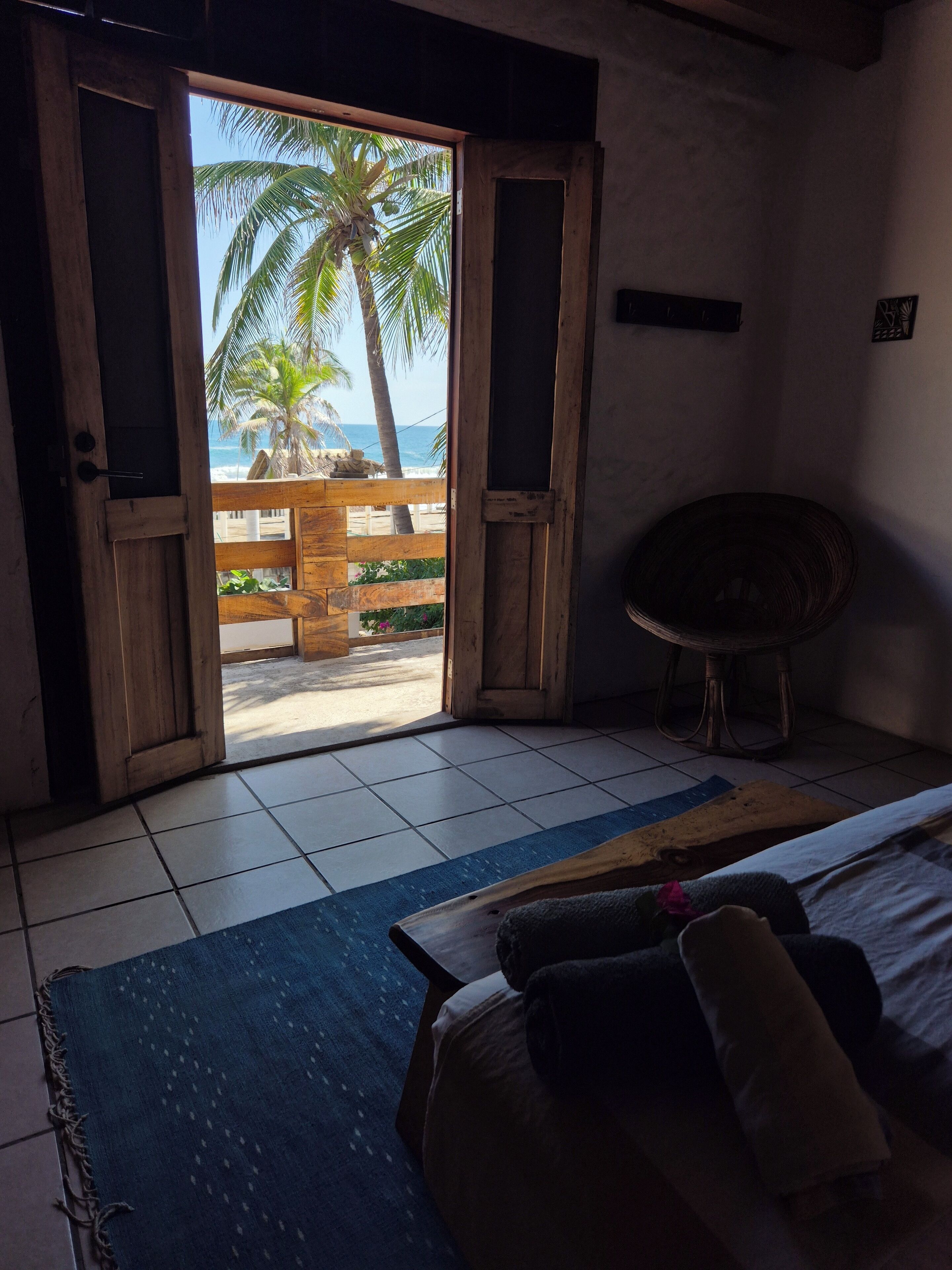 Quarto com uma cama, porta de madeira que dá para uma varanda com vista para palmeiras e o mar.