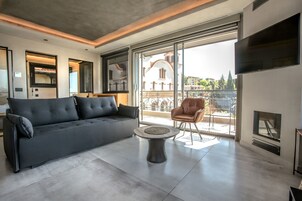 Uma sala de estar moderna com um sofá cinzento, uma mesa de centro redonda e uma poltrona de couro. Uma lareira é visível à direita, e uma grande janela oferece vista para edifícios lá fora.