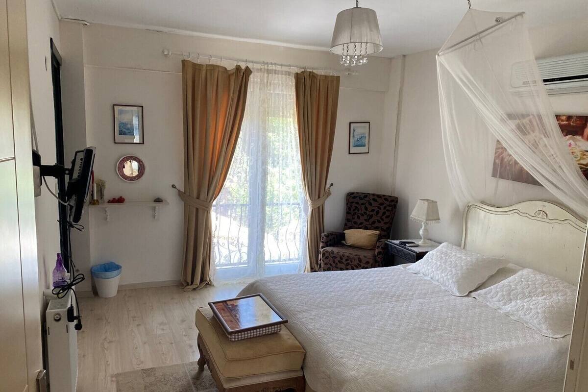 Quarto com cama, cadeira, mesa de cabeceira, luminária, janela com cortinas e iluminação no teto.