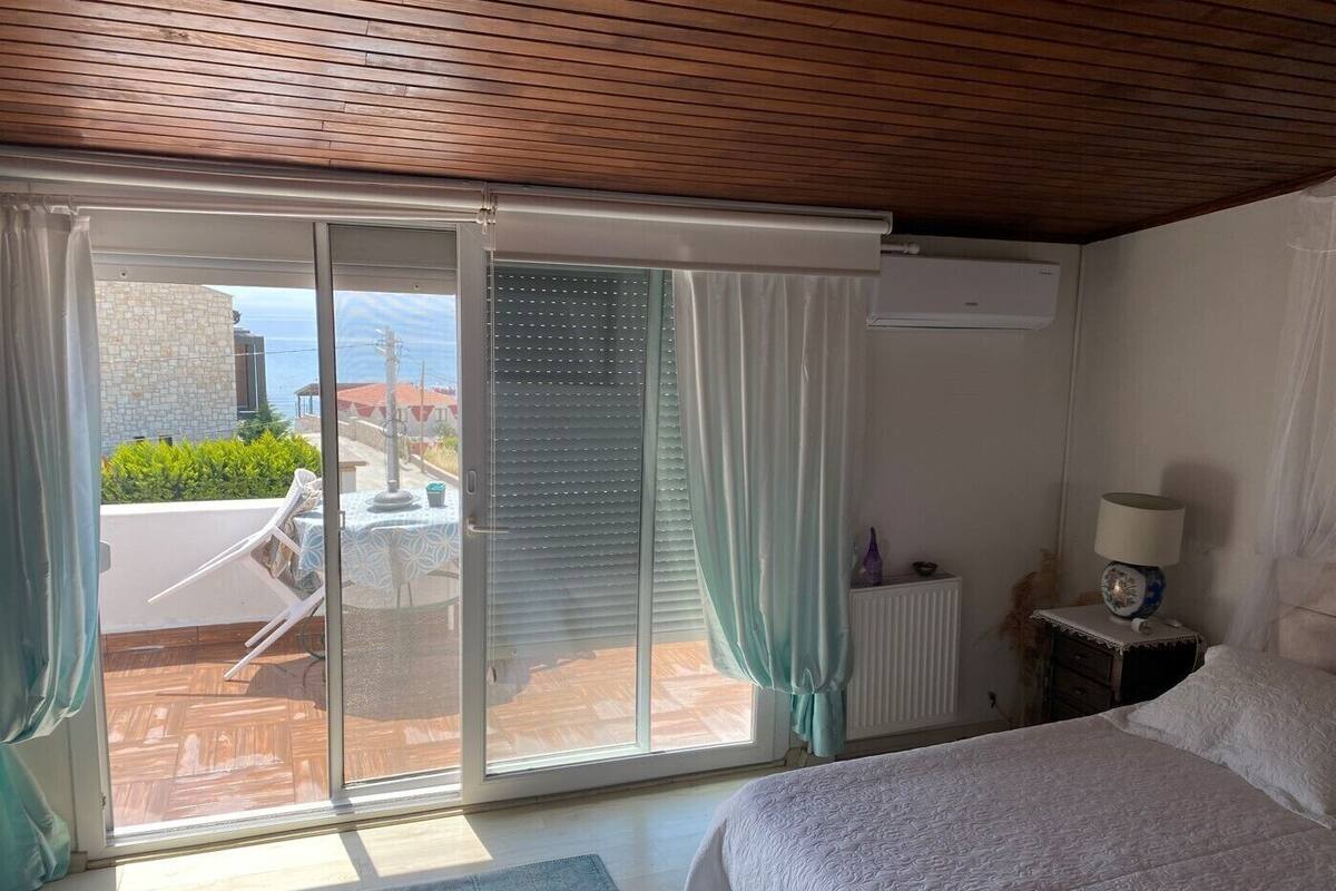 Quarto com uma grande porta de vidro deslizante que dá para uma varanda, uma cama com colcha branca, uma mesa de cabeceira com abajur e um radiador.