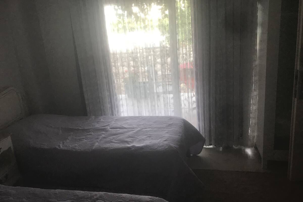 Um quarto com pouca luz, uma cama e uma janela coberta por cortinas finas.