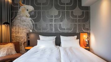 Quarto de hotel com um grande mural na parede retratando um cacatuá, uma cama com roupa de cama branca, uma pequena mesa redonda e um sofá com um travesseiro macio.