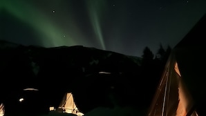 Uma tenda com estrutura de vidro, situada diante de uma paisagem nevada sob um céu noturno com a aurora boreal.
