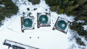 Vista aérea de um resort com três piscinas de telhado verde, rodeado por neve, situado numa área arborizada.