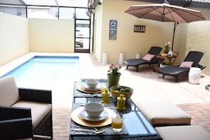 Uma área à beira da piscina com mesa posta para refeições, cadeiras de descanso e guarda-sol.