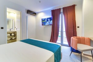 Quarto de hotel com cama, televisão, poltrona e banheiro com espelho e porta-toalhas.