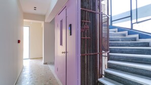 Um corredor com elevador roxo, escada com corrimão de metal e uma janela grande que traz luz natural.
