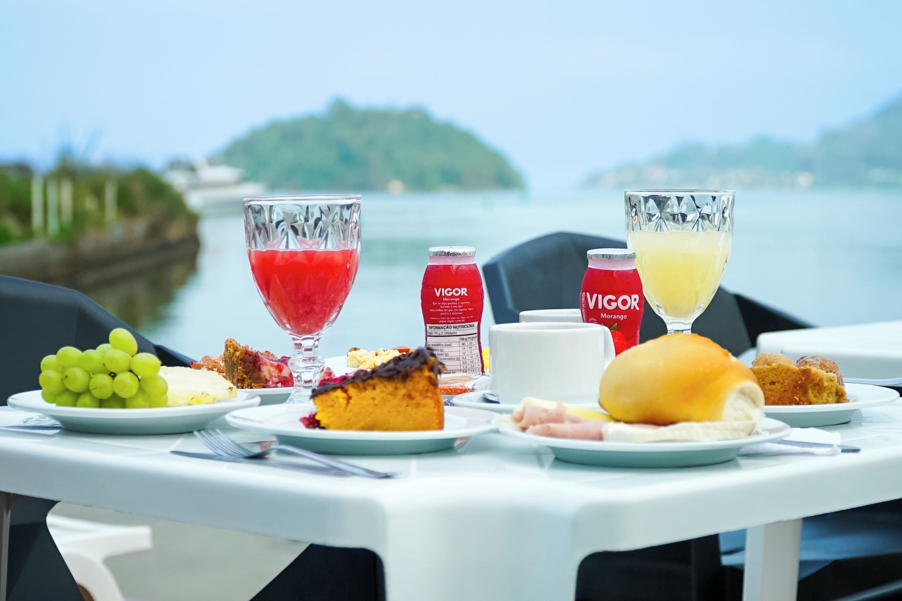 Uma mesa com comida e bebida, vista panorâmica da água e vegetação, e uma garrafa de suco Vigor Morango.