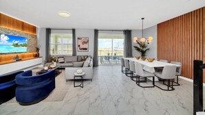 Uma sala de estar moderna com uma televisão grande, assentos confortáveis, um espaço de jantar com mesa e cadeiras, e vista para o exterior.