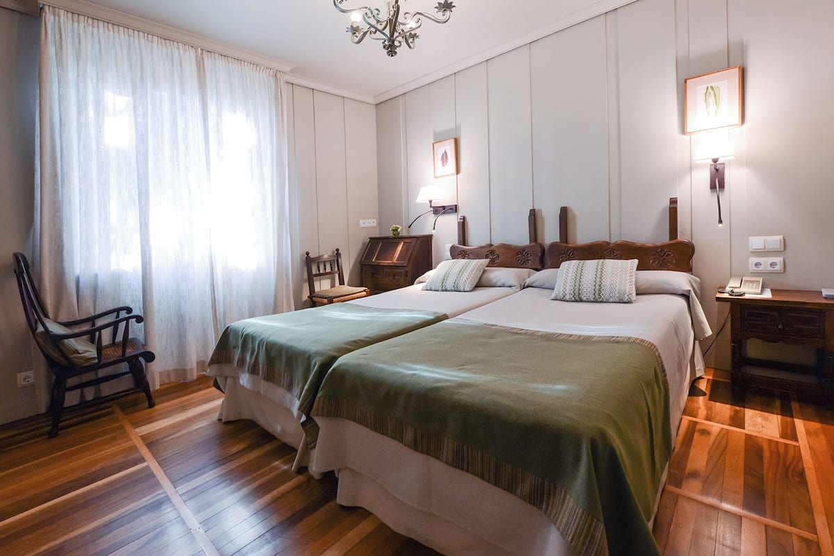 Quarto de hotel com uma cama grande, duas mesas de cabeceira, uma cadeira, uma escrivaninha e um candelabro.