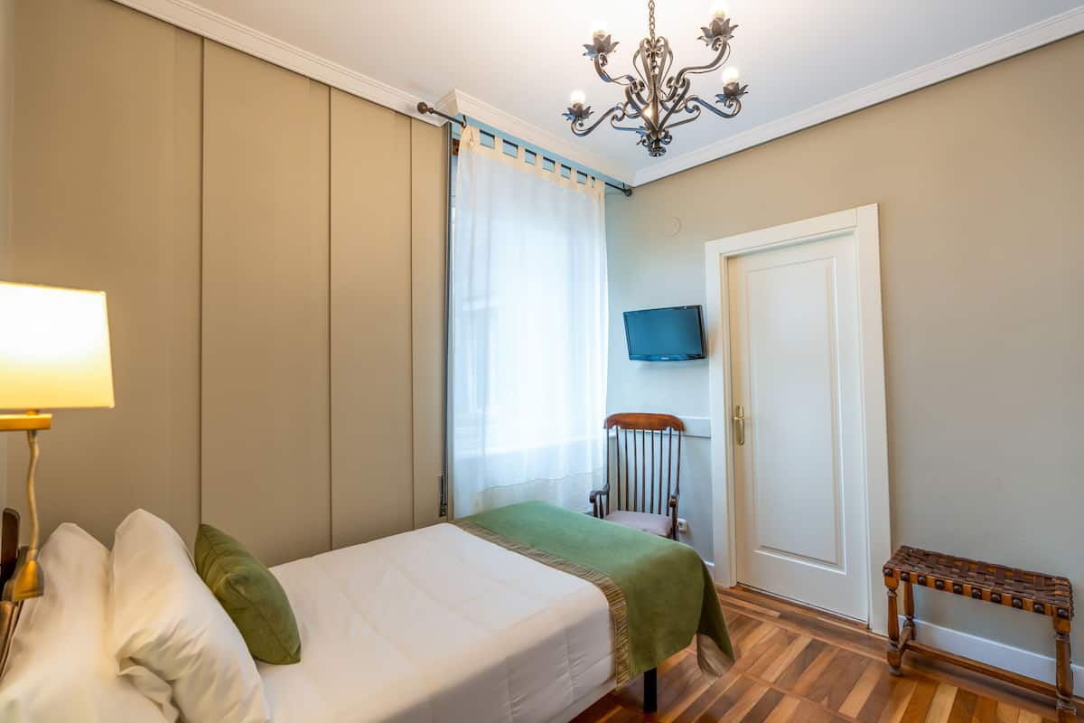 Quarto com cama, luminária de cabeceira, manta verde, banco de madeira, televisão e porta.