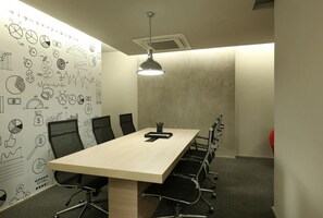 Uma sala de conferência com uma mesa comprida, cadeiras pretas, uma luminária no teto e uma parede com diversos gráficos e diagramas.