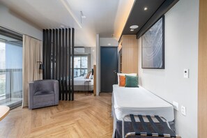 Quarto de hotel moderno com uma cama, uma poltrona, uma mesinha e vista da cidade através de grandes janelas.