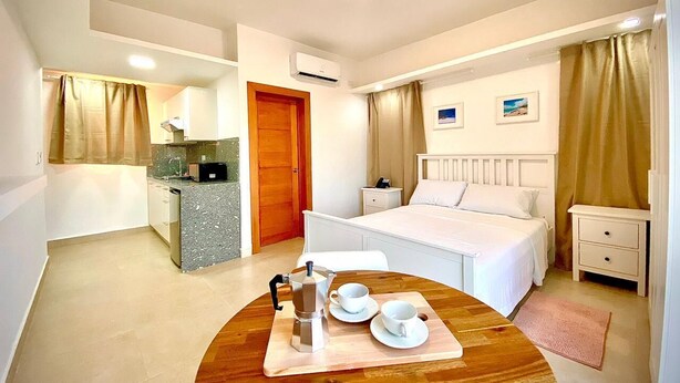 Quarto de hotel com cama, mesa de cabeceira, pequena escrivaninha, cozinha compacta e área de jantar posta para o café da manhã.