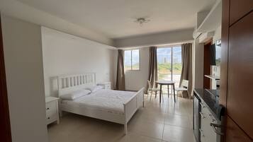 Quarto com cama, mesas de cabeceira, uma pequena mesa com cadeiras, ventoinha no teto e vista através de grandes janelas.