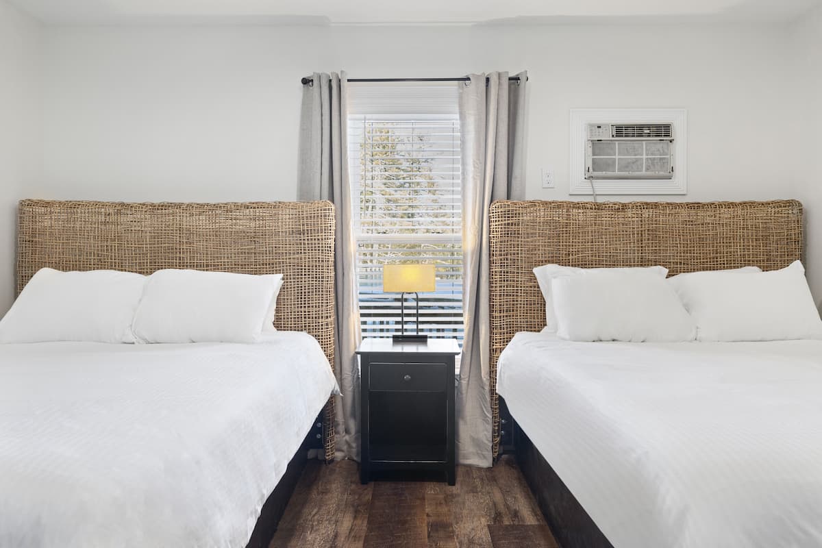 Quarto de hotel com duas camas, uma mesa de cabeceira, uma luminária e um aparelho de ar condicionado.