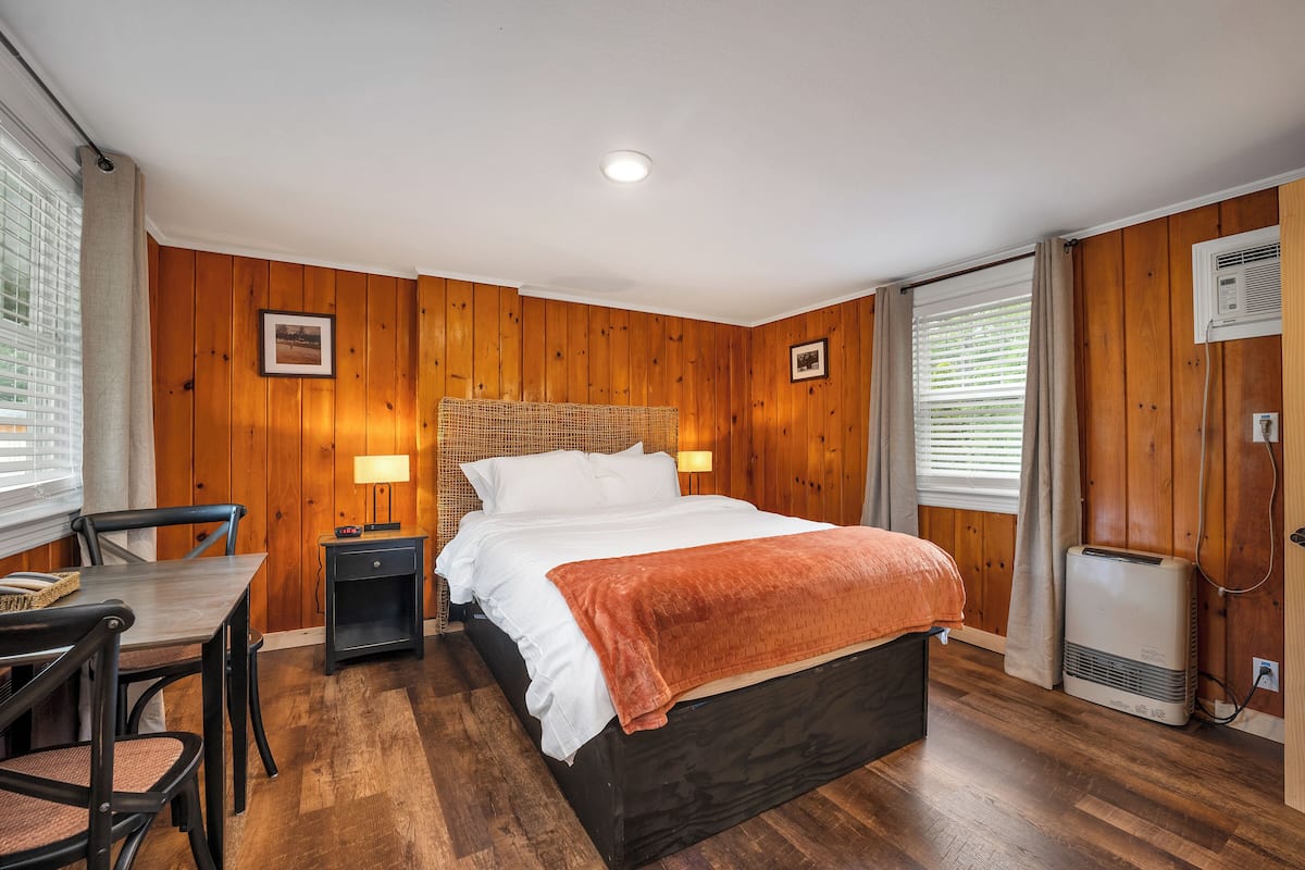 Quarto com cama, mesa de cabeceira, luminária e janela com persianas.