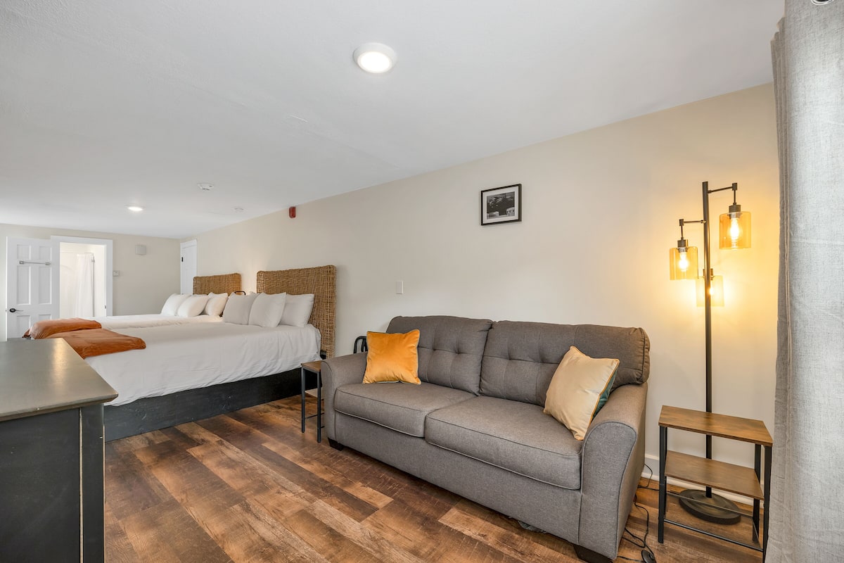 Quarto moderno com cama, sofá cinza, mesinha e abajur.