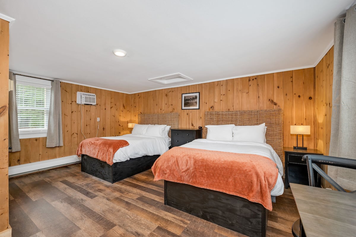 Quarto com duas camas, paredes de madeira, janela com persianas, uma mesa e uma cadeira.
