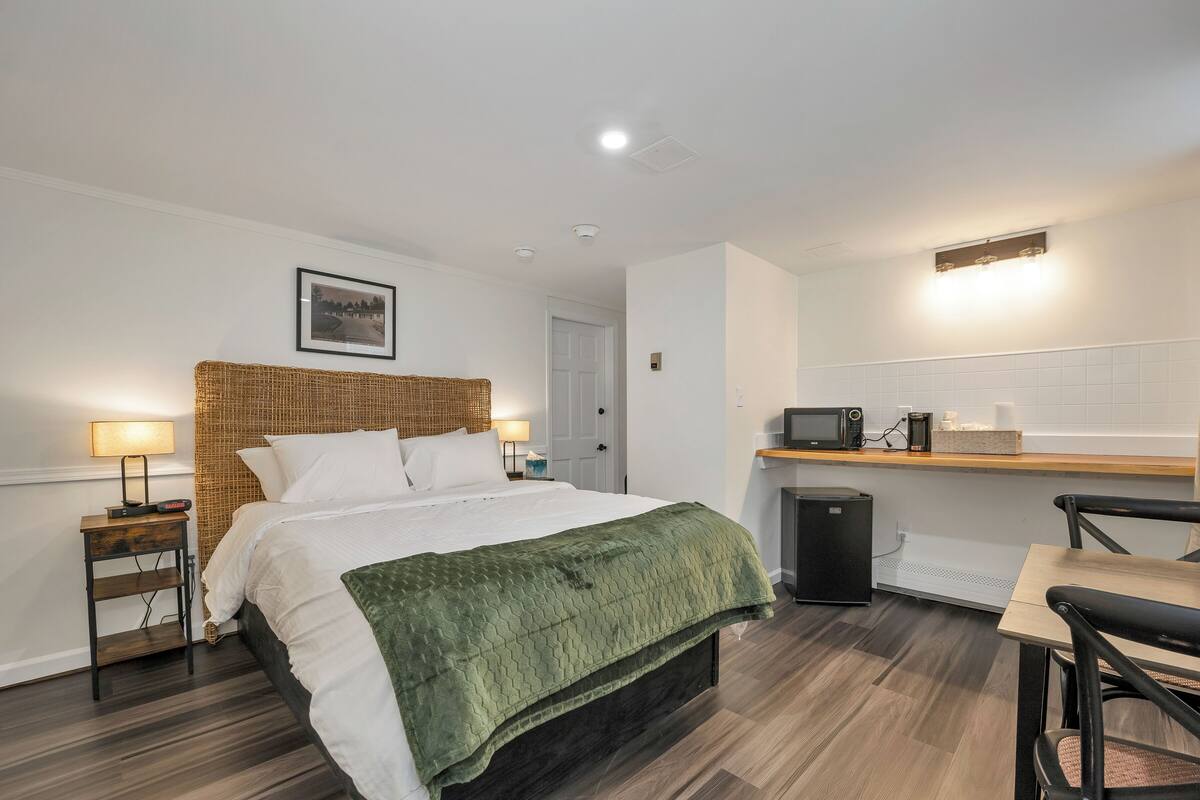 Quarto de hotel com cama, mesa de cabeceira, luminária e uma área de cozinha compacta com micro-ondas e torradeira.