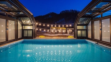 Piscina com estrutura de vidro, rodeada por um deck com luzes de corda e vista para uma paisagem montanhosa.